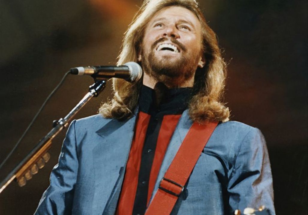 Under the soft glow of a stage light, Barry Gibb stands alone, guitar ...
